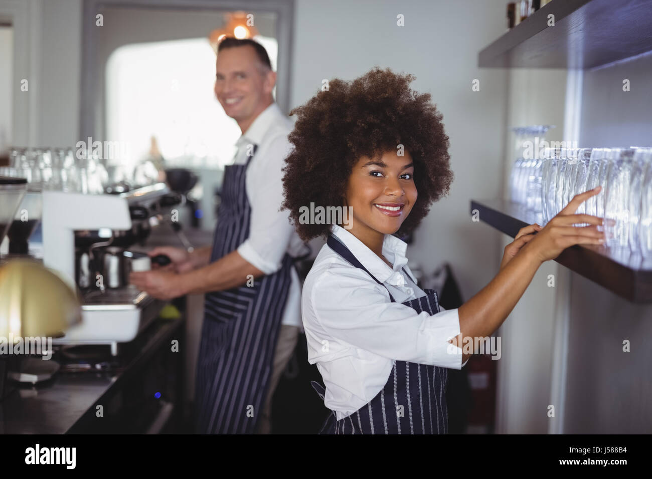 Portrait of happy waitress arranging glass in shelf at counter of ...