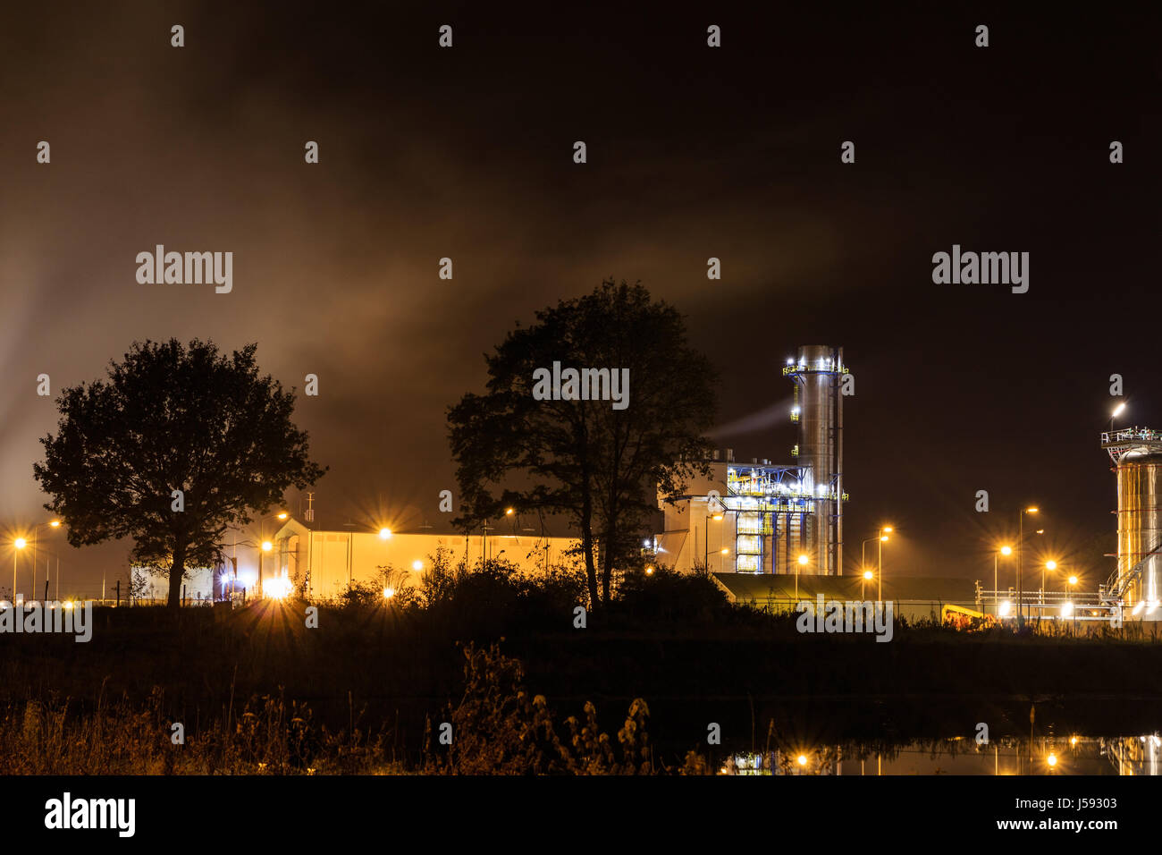 Petrochemical plant at night, trees and a pond in the foreground ...