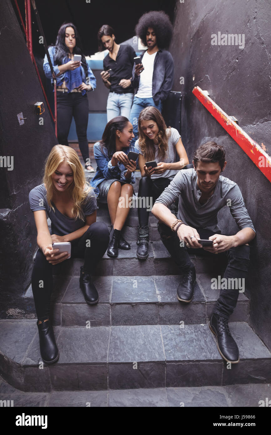 Young friends using mobile phones while sitting on steps at nightclub ...