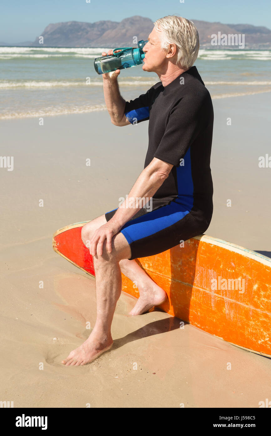 Senior man drinking water while sitting on surfboard at beach Stock ...