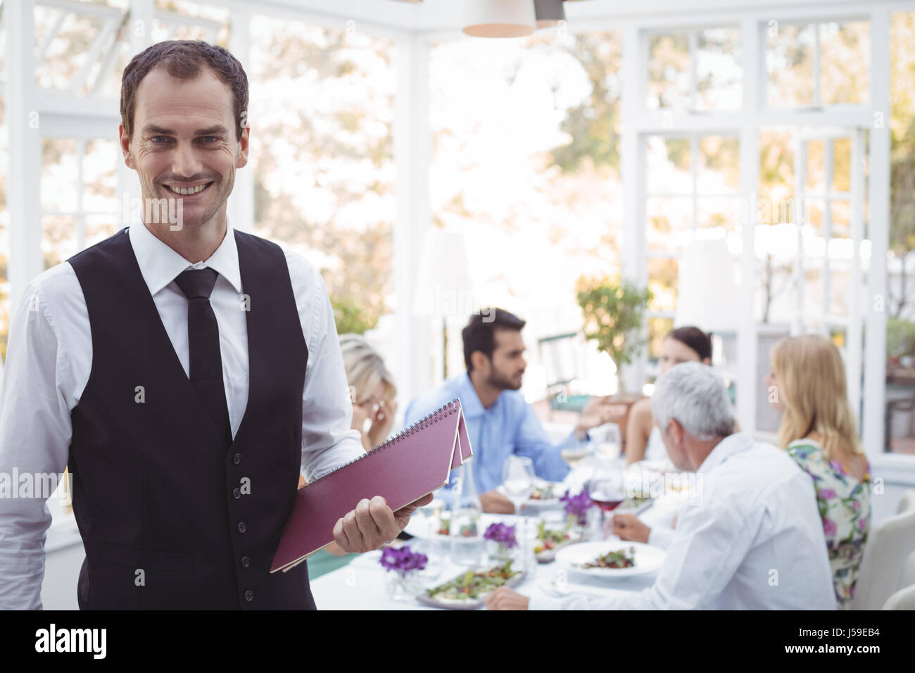 Portrait of smiling waiter holding menu while friends dining in ...