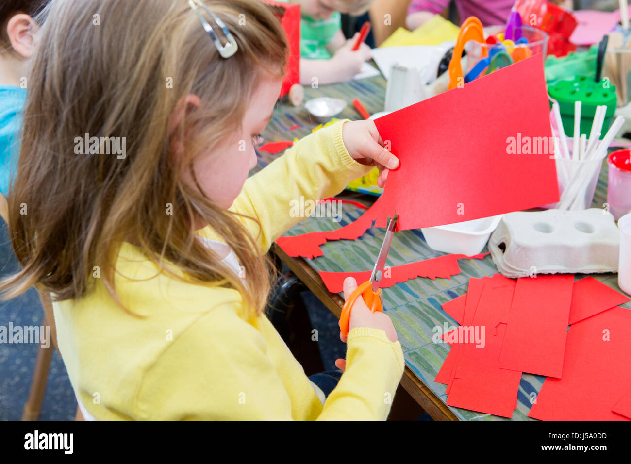Little girl using scissors to cut shapes from a red piece of paper. she ...