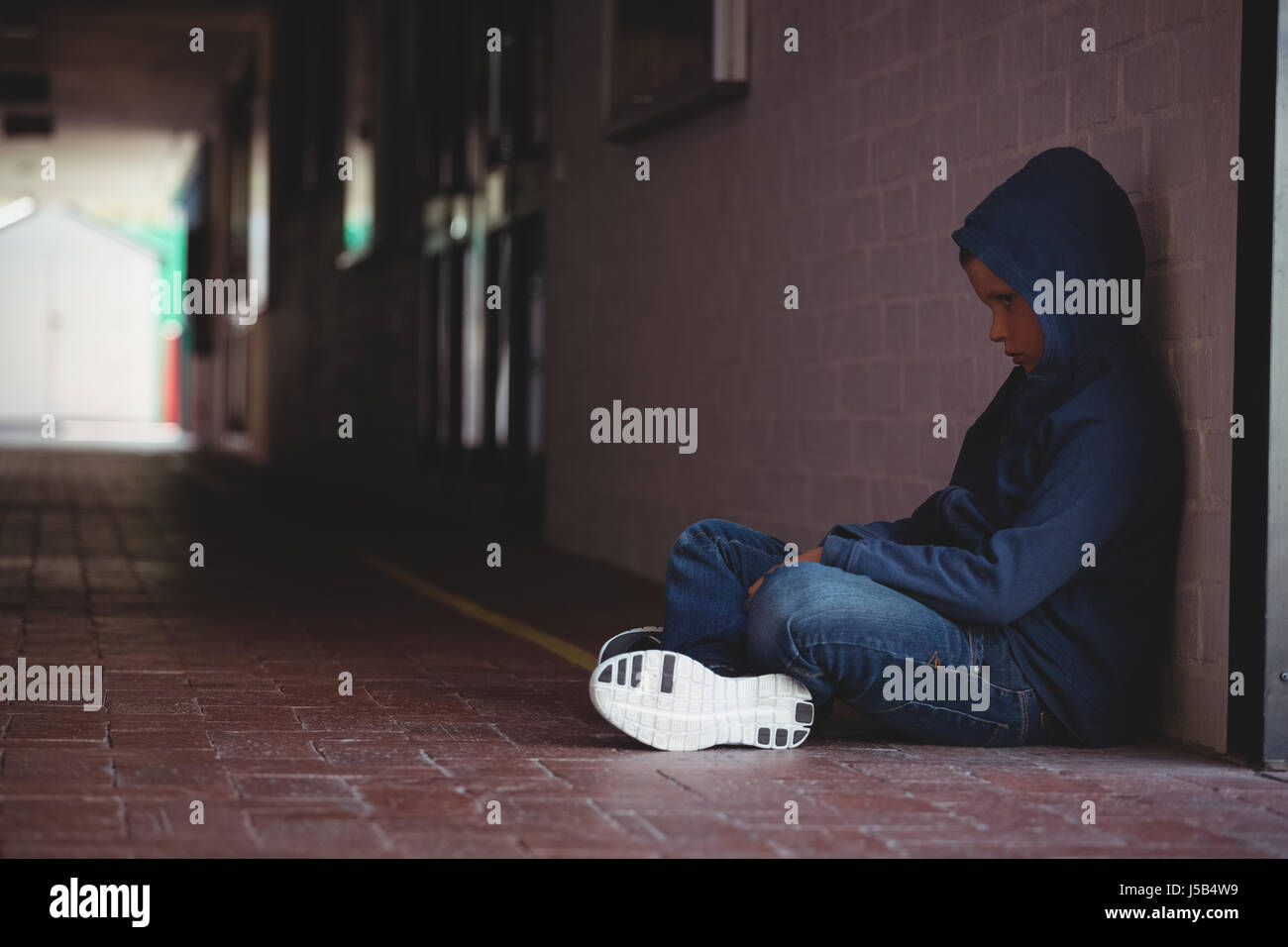 Full length of sad boy sitting on pavement by wall in corridor at ...