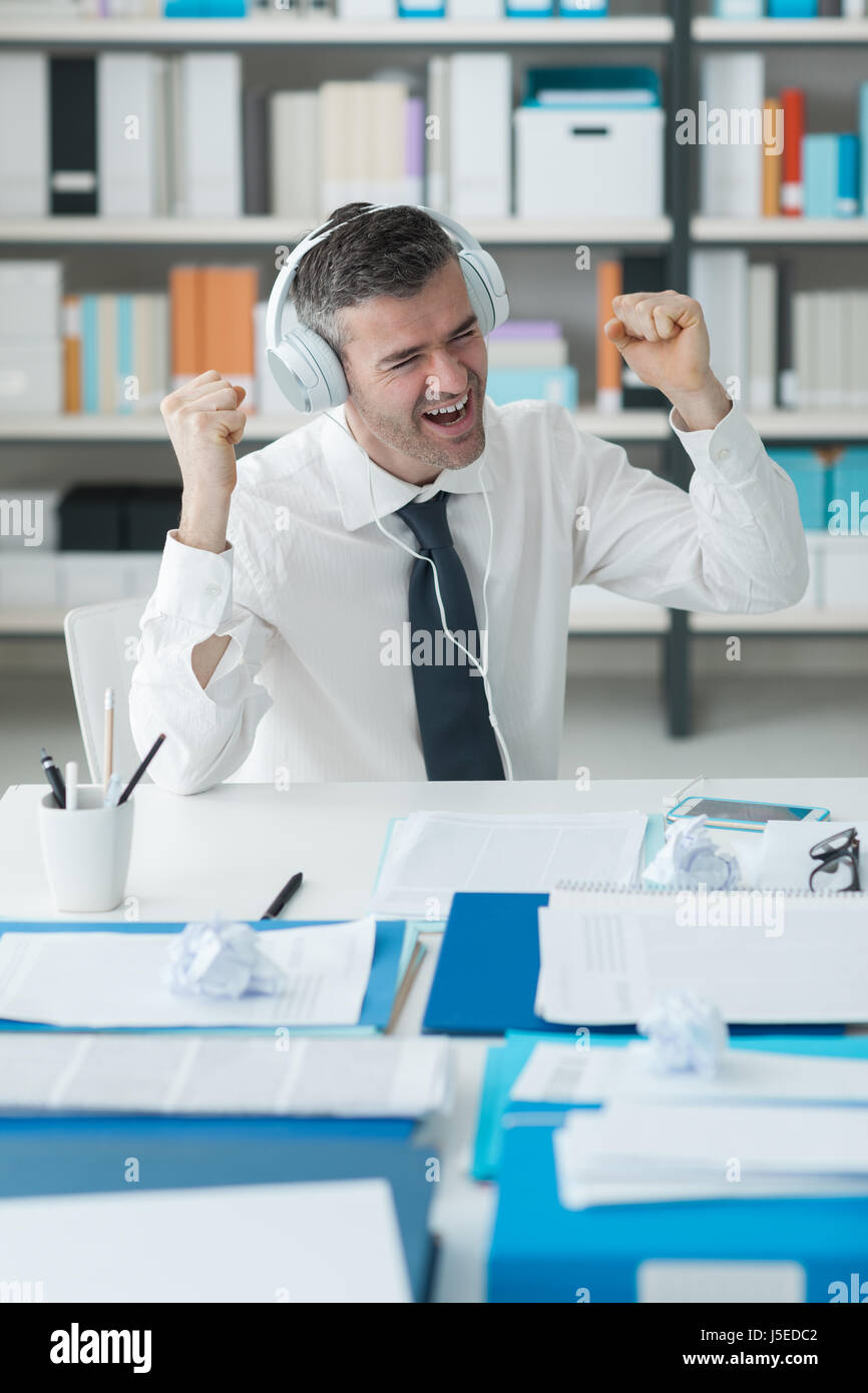Cheerful businessman sitting at office desk, listening to music using ...