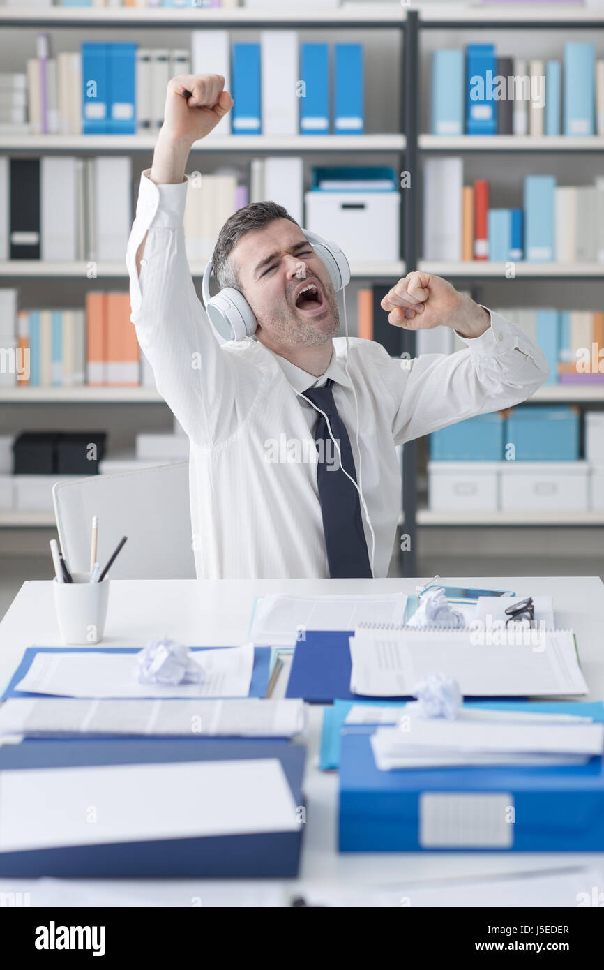 Cheerful businessman sitting at office desk, listening to music using ...