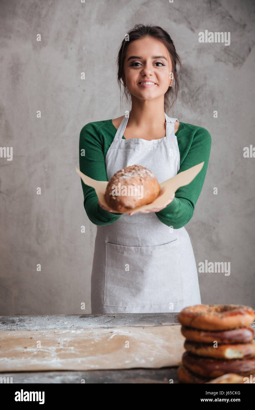 Image of young happy lady baker standing and holding bread. Looking at ...