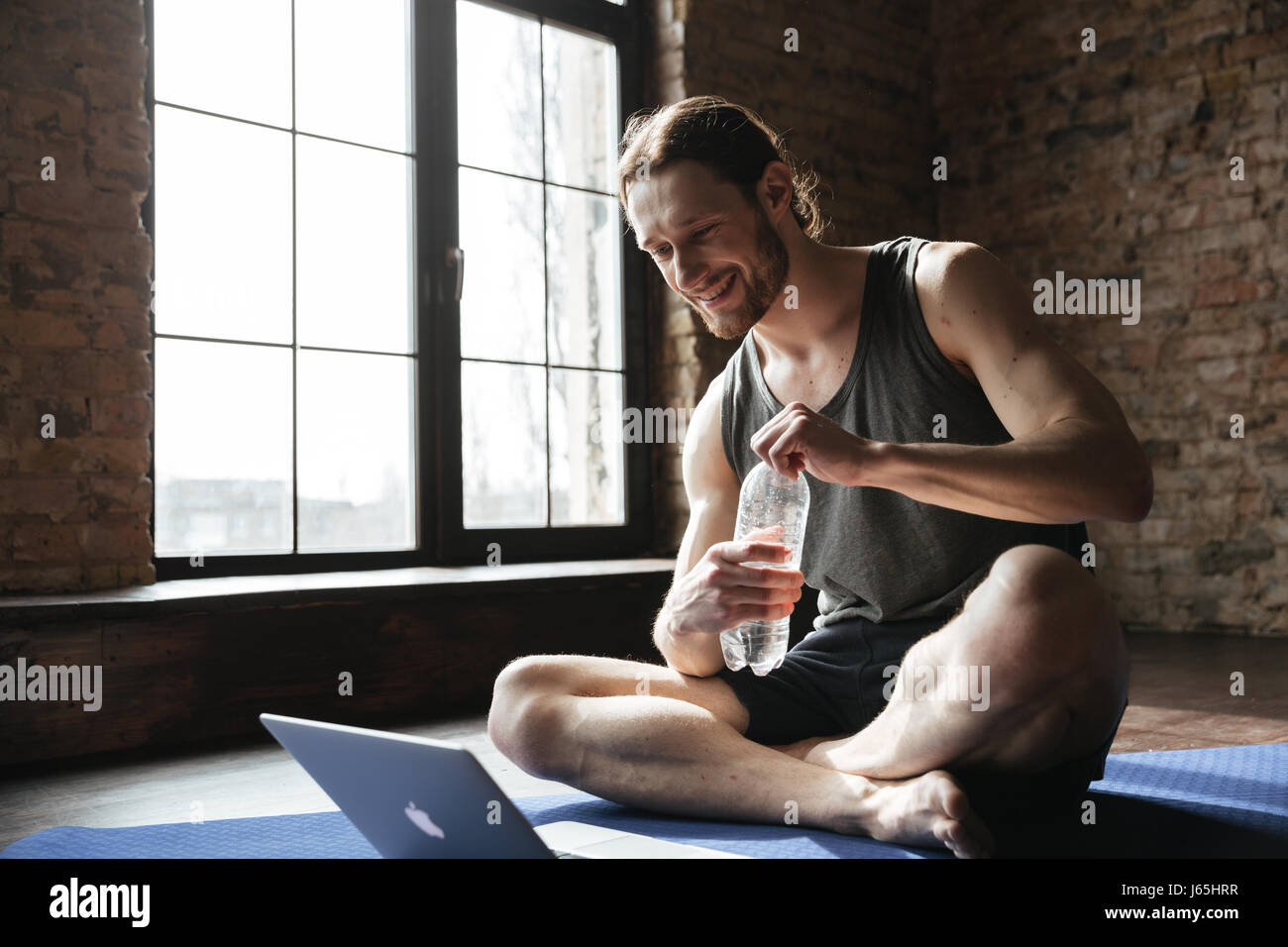 Picture of happy strong sportsman drinking water on floor in gym using ...