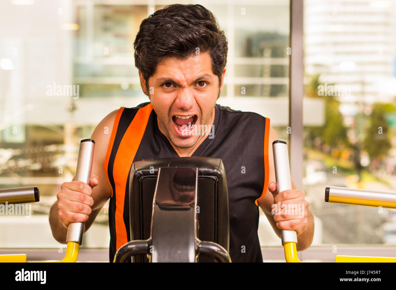 Handsome young man working on fitness machine at gym and screaming at ...