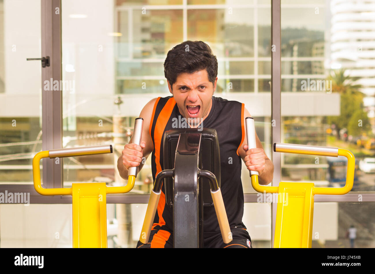 Handsome young man working on fitness machine at gym and screaming at ...