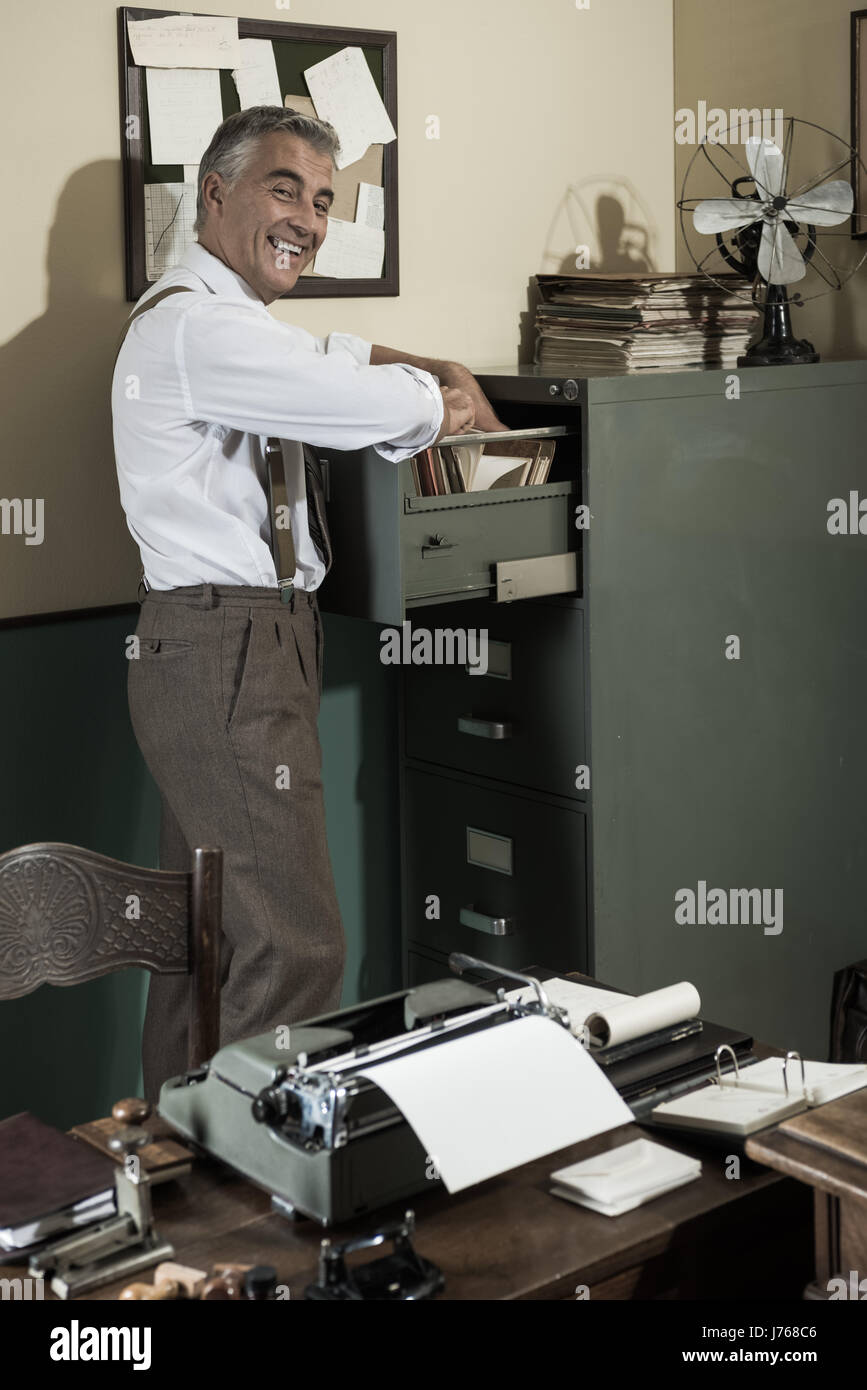 Cheerful smiling employee in 1950s style office searching for a file in ...