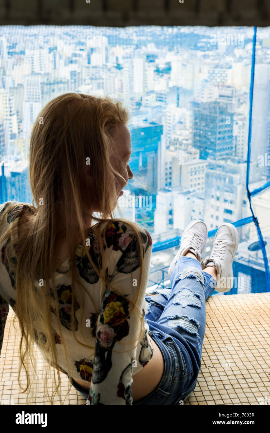 Young woman in high rise building sitting in window, looking over city ...