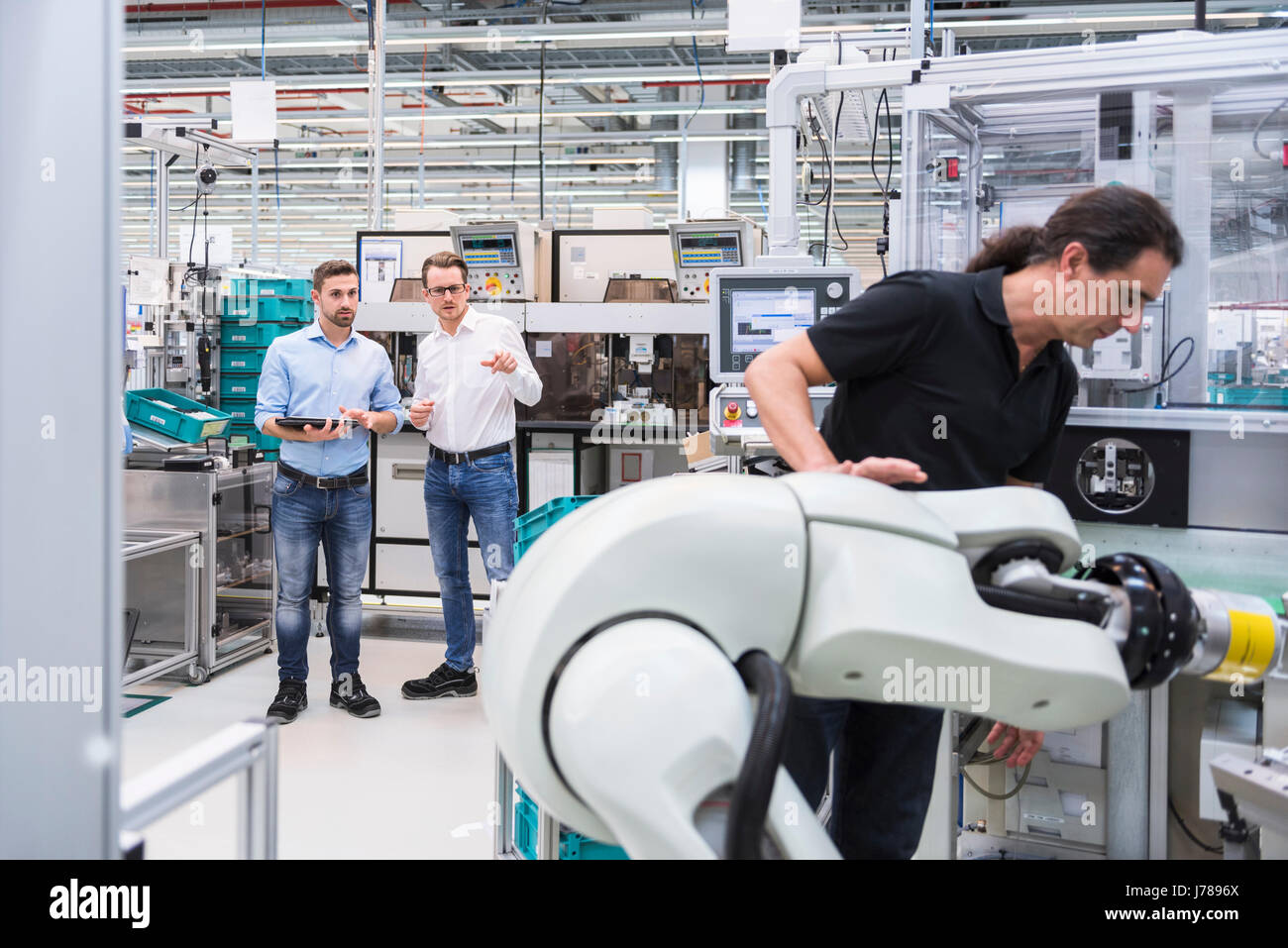 Man operating assembly robot in factory with two men in background ...