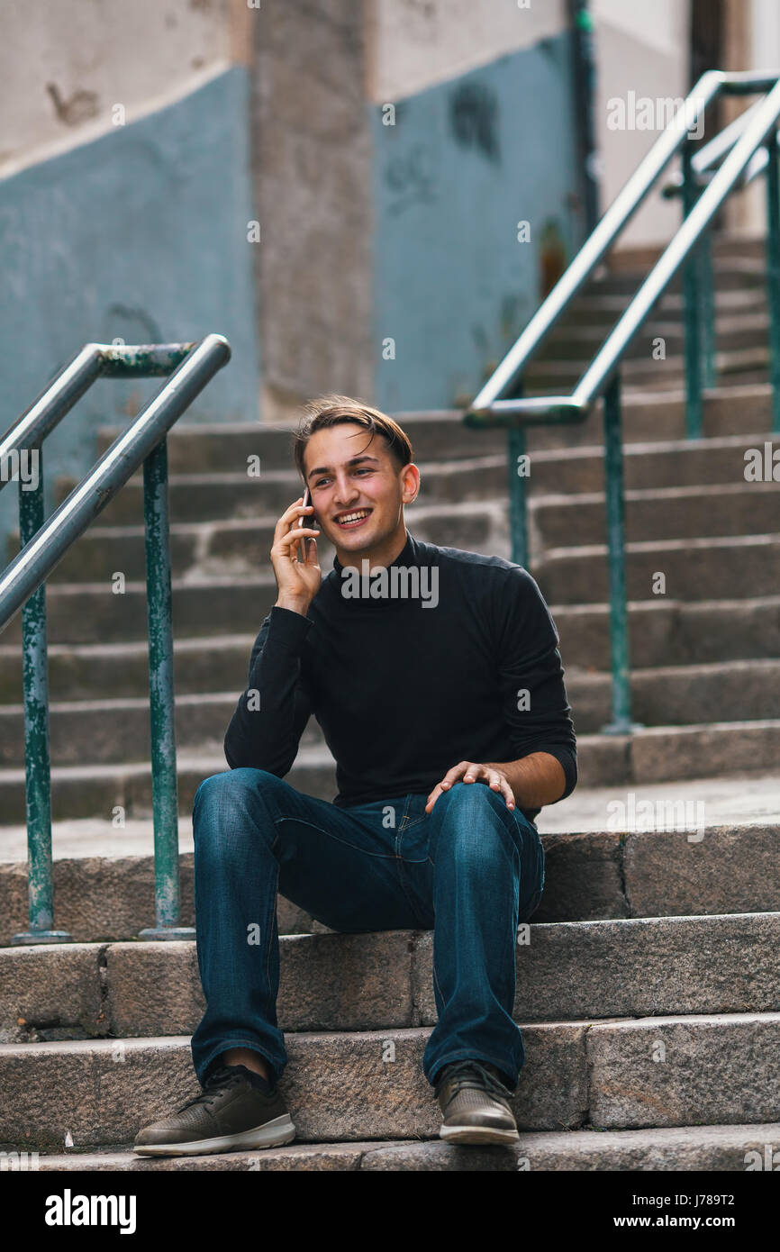 Handsome young man talking on a mobile sitting on the steps of outdoors ...