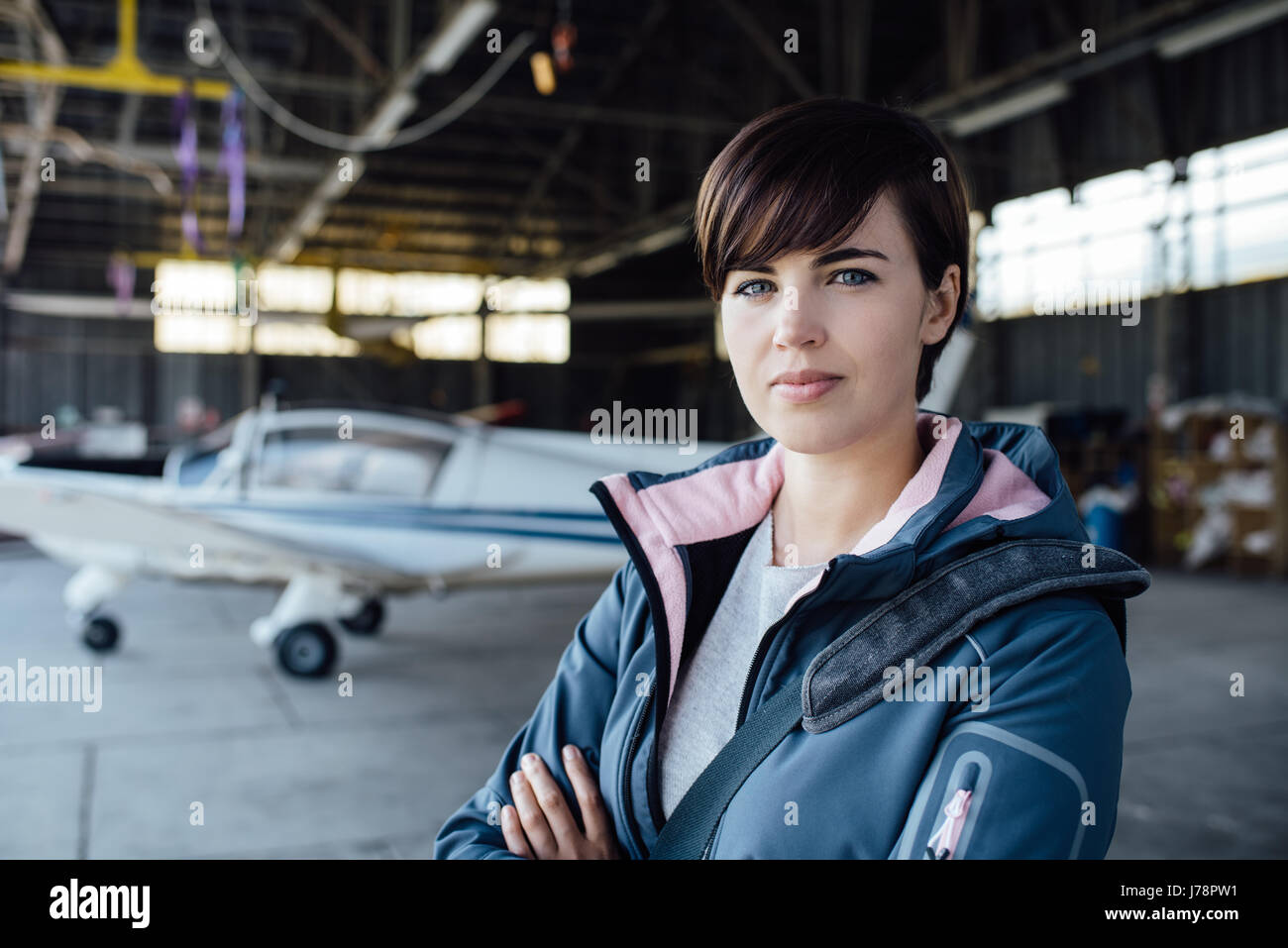 Confident young female pilot posing in the hangar with arms crossed ...
