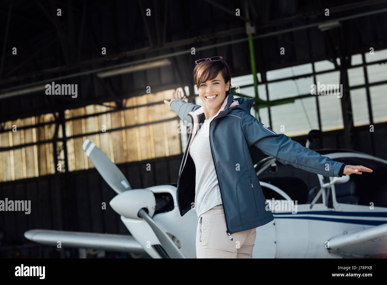 Smiling cheerful female pilot posing in the hangar with arms raised ...