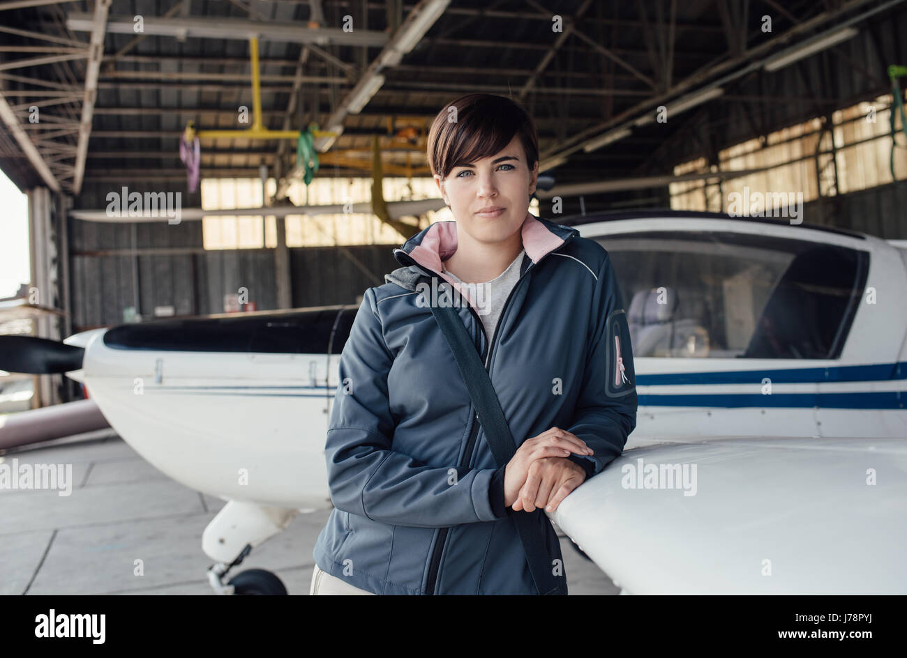 Young smiling female pilot posing in the airport hangar, she is leaning ...