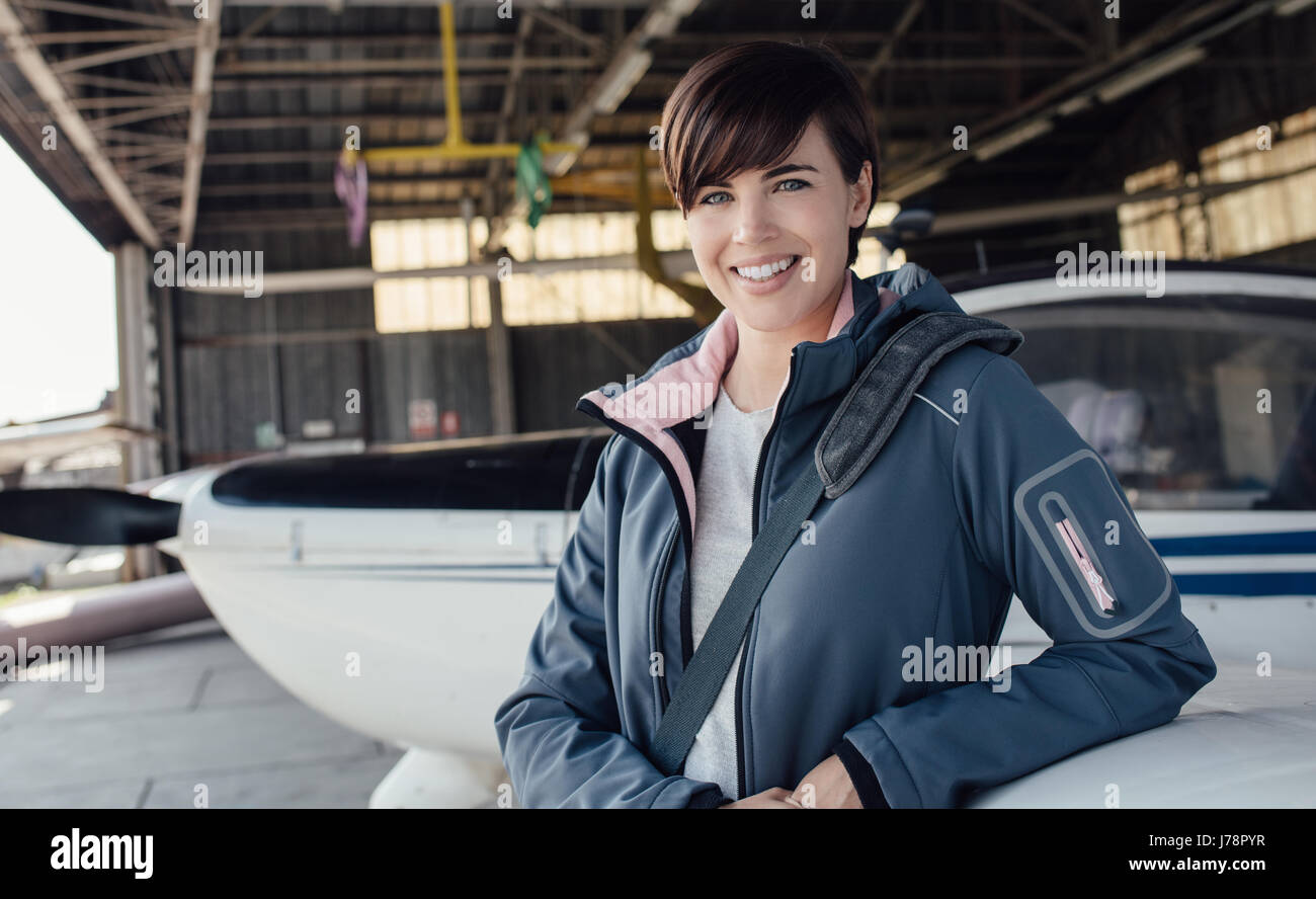 Young smiling female pilot posing in the airport hangar, she is leaning ...