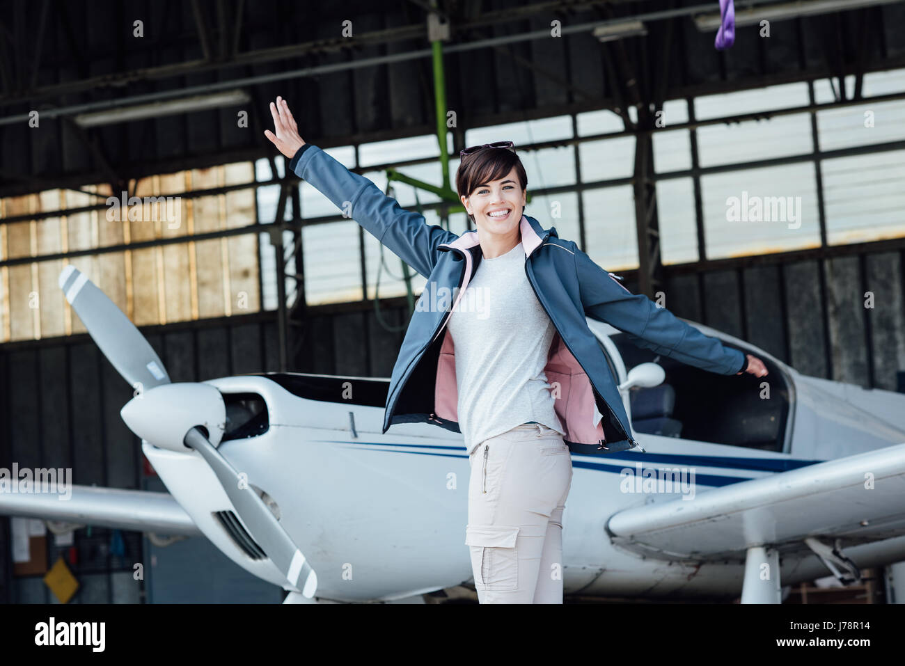 Smiling cheerful female pilot posing in the hangar with arms raised ...