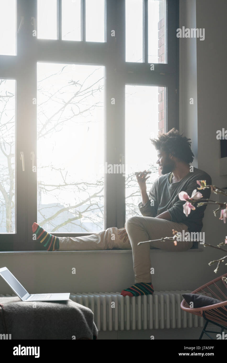 Man sitting on window sill in living room looking outside talking into ...