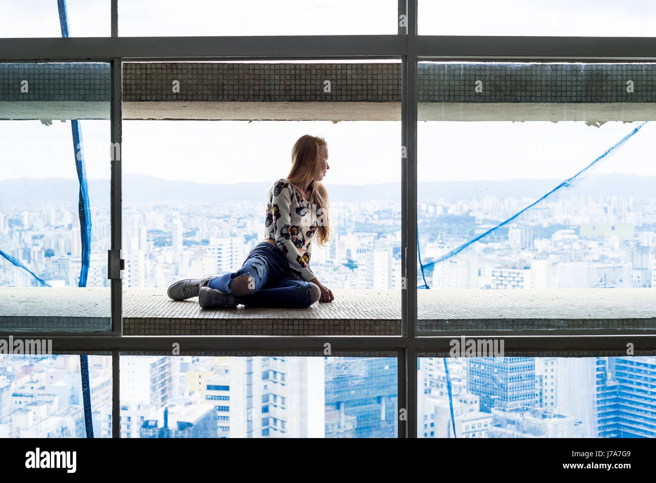 Young woman in high rise building sitting in window, looking over city ...