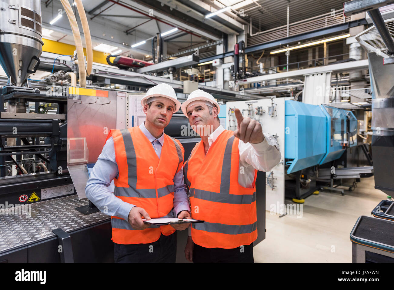 Two men wearing safety vests and hard hats talking in factory shop ...