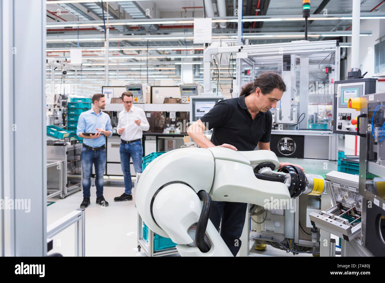Man operating assembly robot in factory with two men in background ...