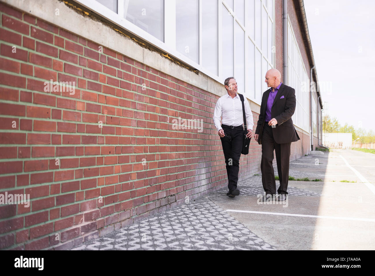 Two men walking along factory building talking Stock Photo - Alamy