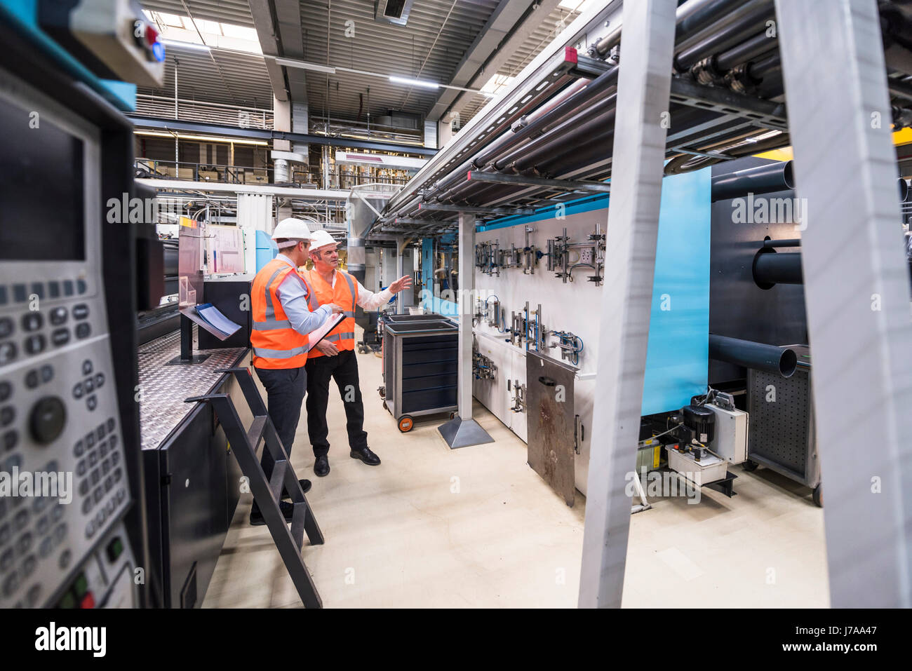 Two men wearing safety vests and hard hats talking in factory shop ...