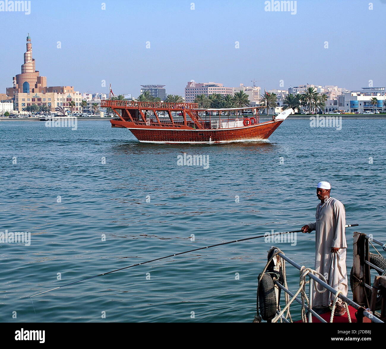 A dhow sailing from the harbour of Doha, Qatar, with a man fishing from ...