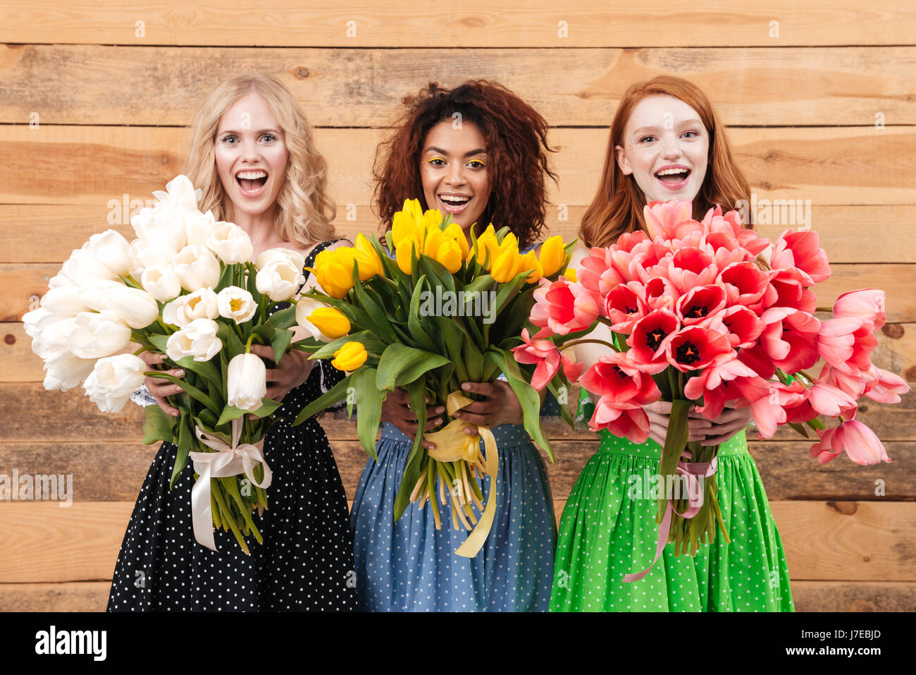 Three happy women in dresses posing near the wooden wall and showing ...