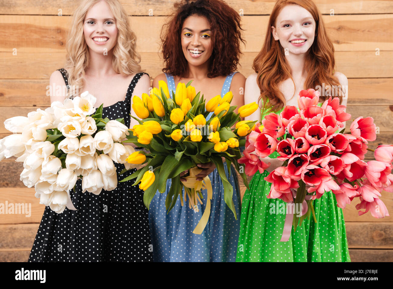 Three happy women in dresses posing near the wooden wall and holding ...