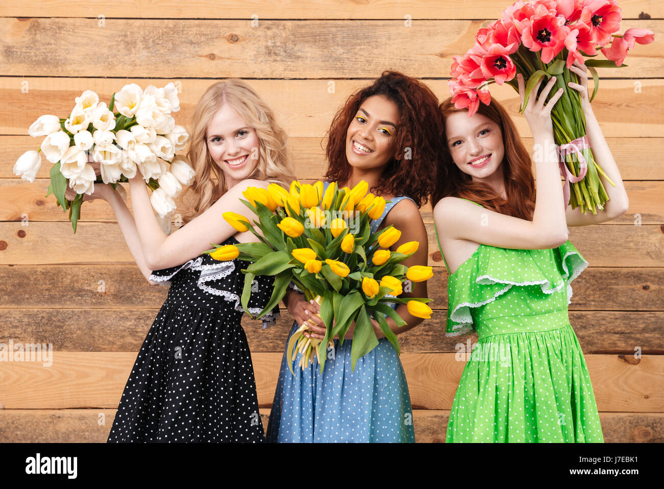 Three pleased women in dresses posing near the wooden wall and holding ...