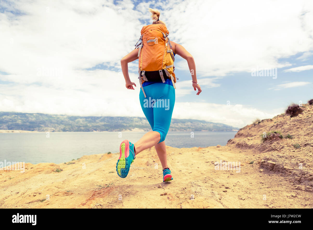Woman running with backpack on rocky trail at seaside and mountains ...