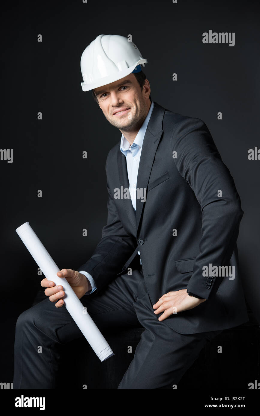 Smiling handsome male engineer in helmet holding blueprint and looking ...