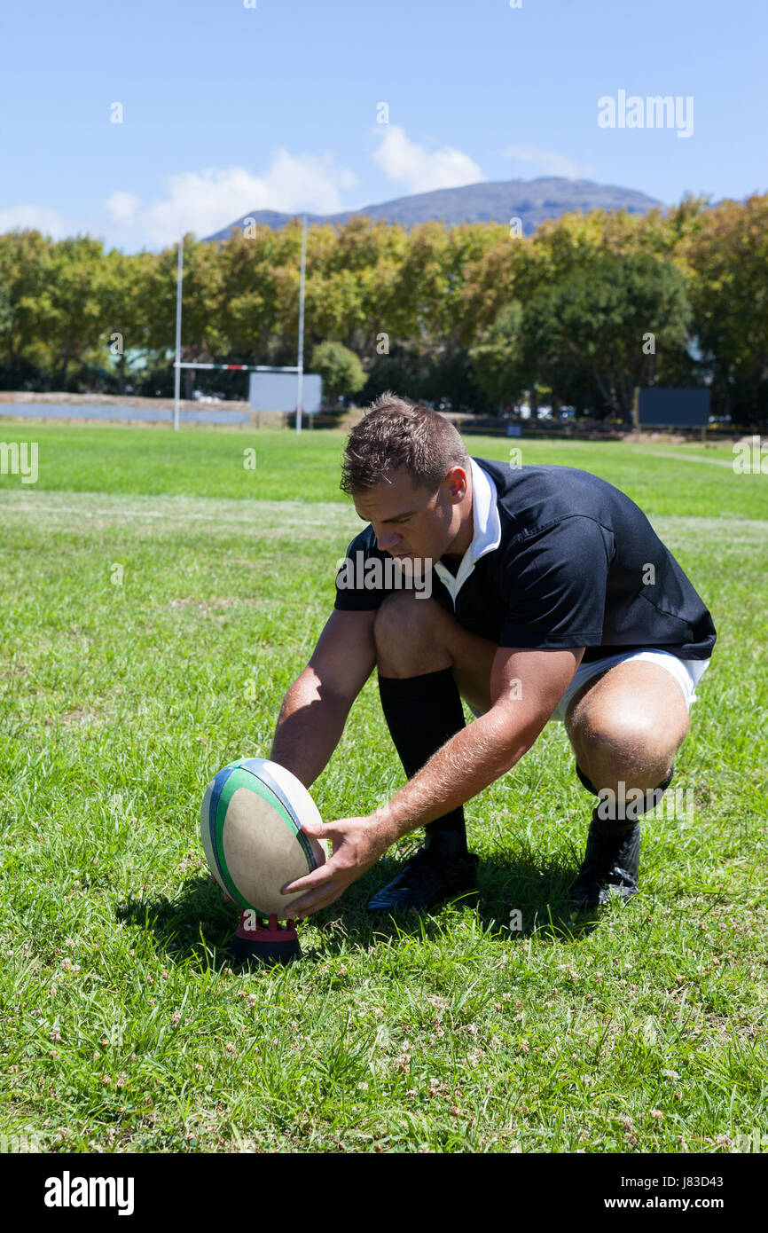 Full length of player kneeling while holding rugby ball on grassy field ...