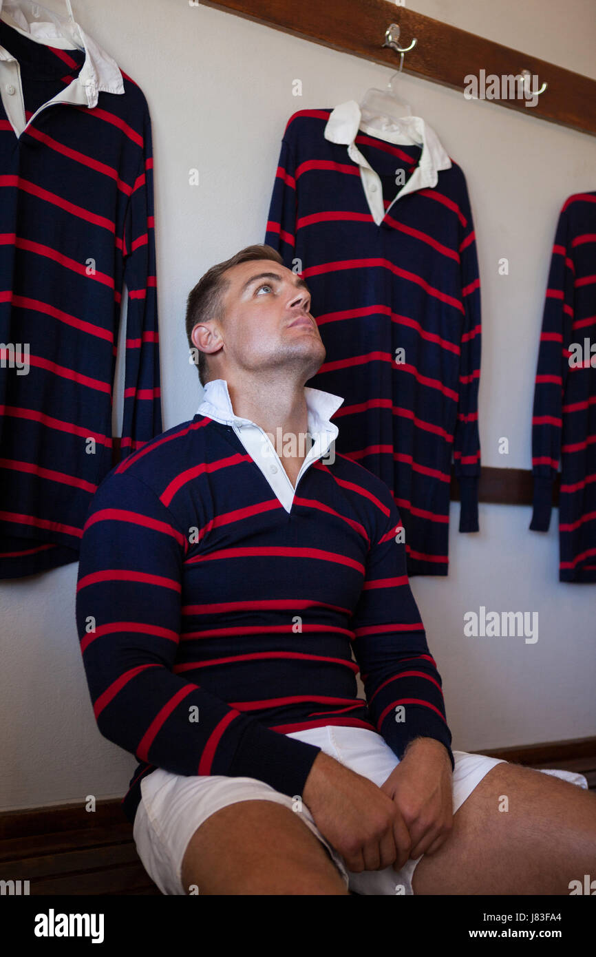 Tired rugby player looking up while sitting on bench at locker room ...