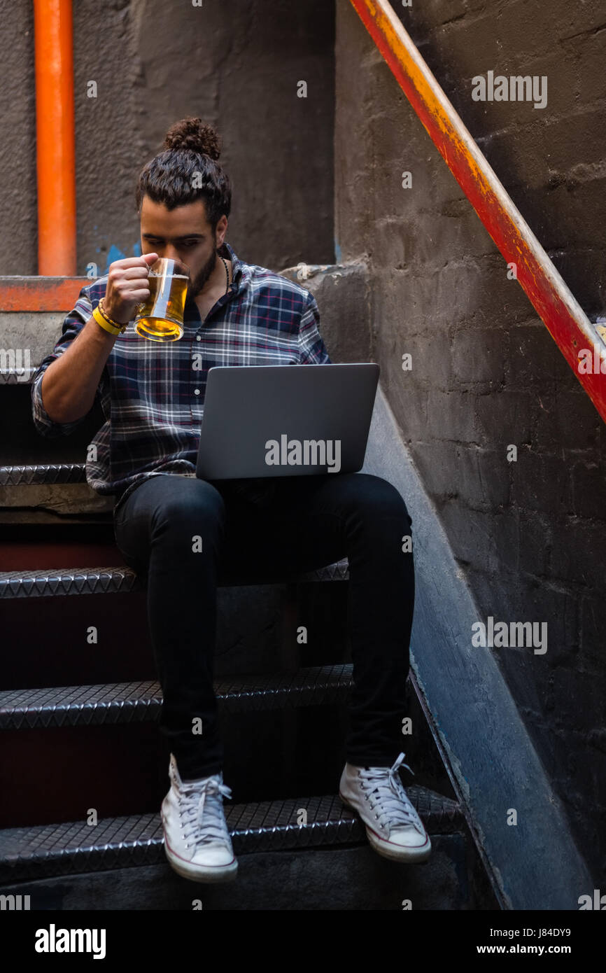 Man using laptop while having beer on staircase of bar Stock Photo - Alamy
