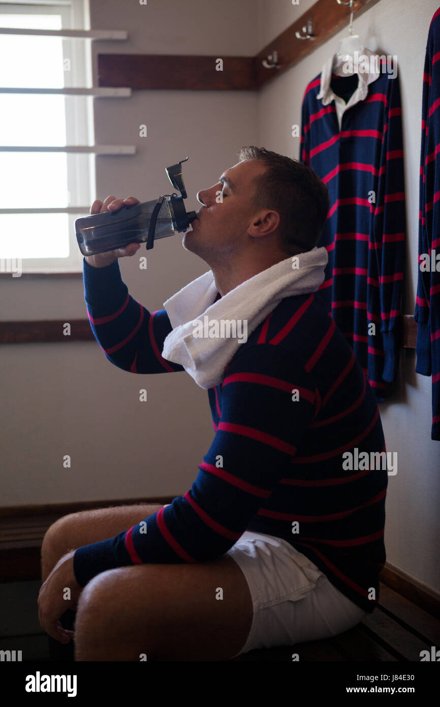 Tired rugby player drinking water while sitting on bench against wall ...