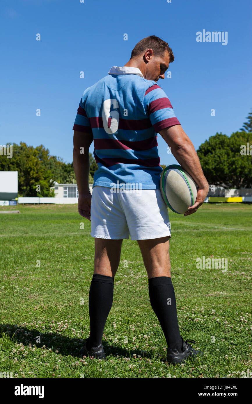 Rear view of rugby player holding ball while standing at playing field ...