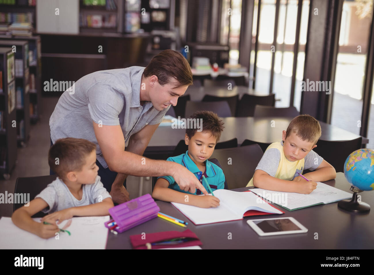 Teacher helping schoolkids with their homework in library at school ...