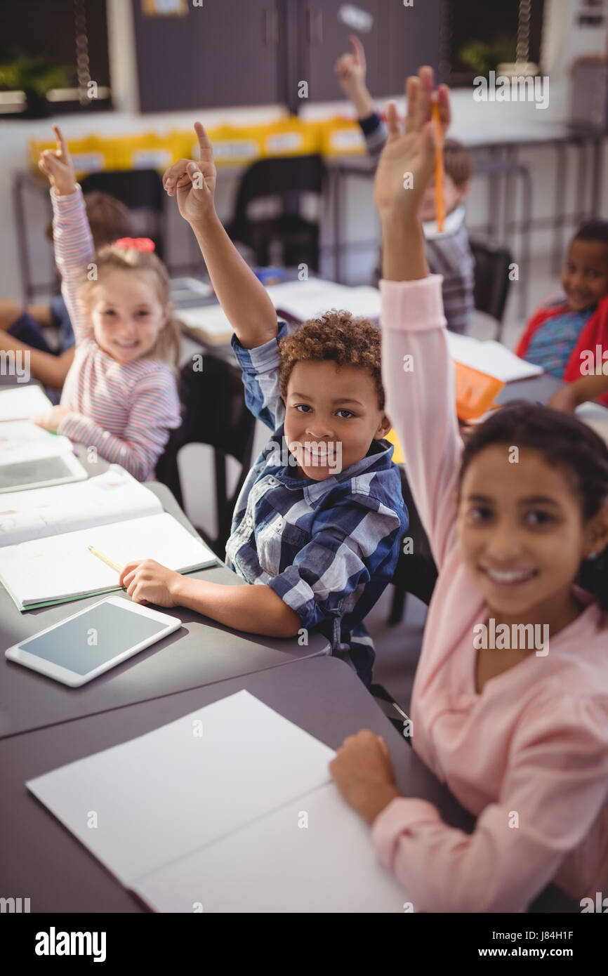 Portrait of happy schoolkids raising their hands in classroom at school ...