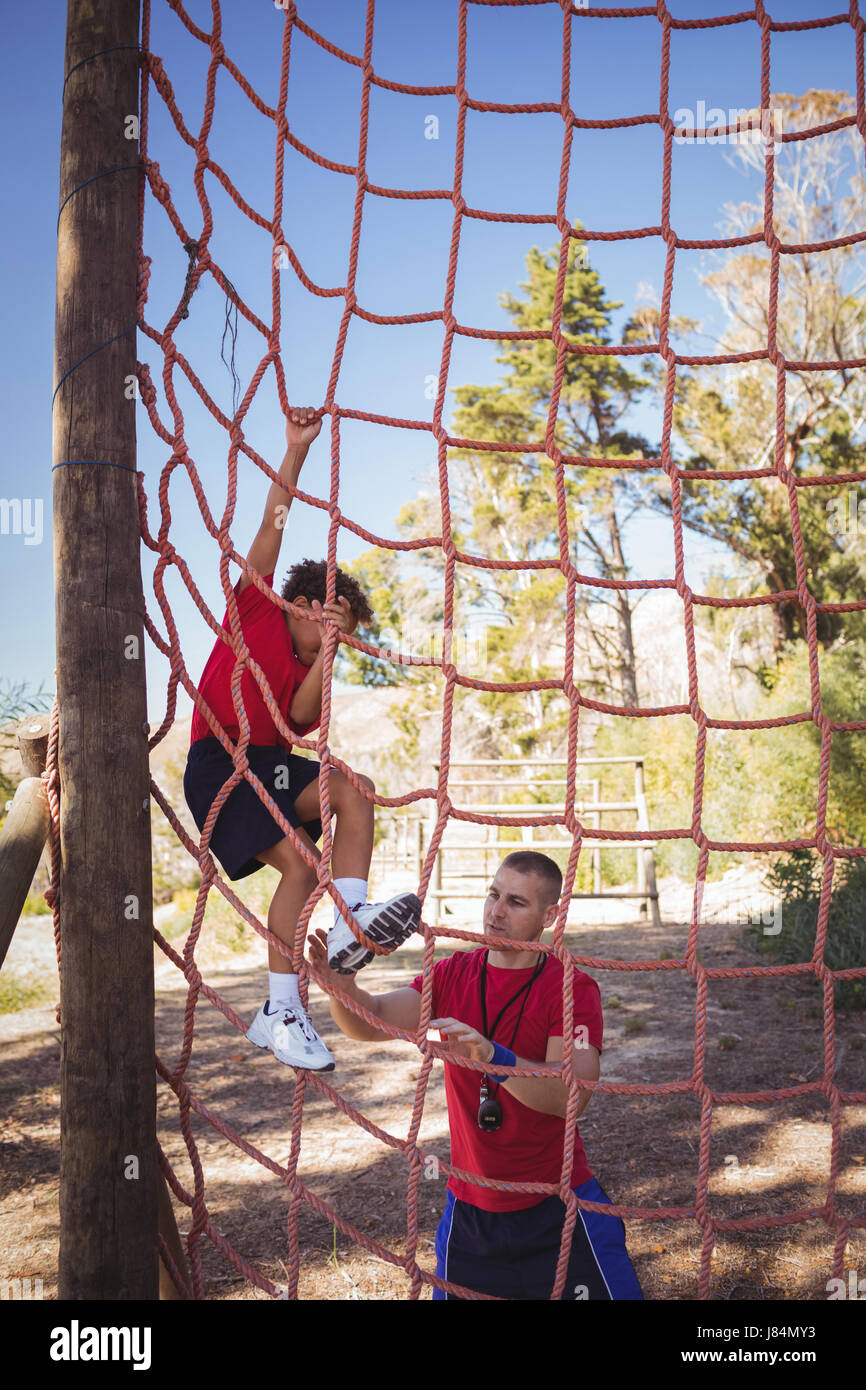 Trainer assisting boy to climb a net during obstacle course training in ...