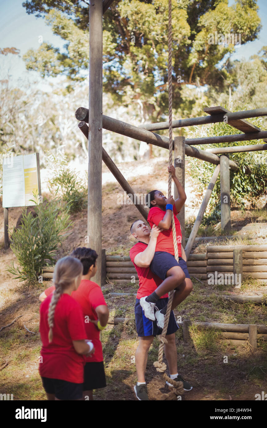 Trainer assisting a kid to climb a rope in the boot camp on a sunny day ...