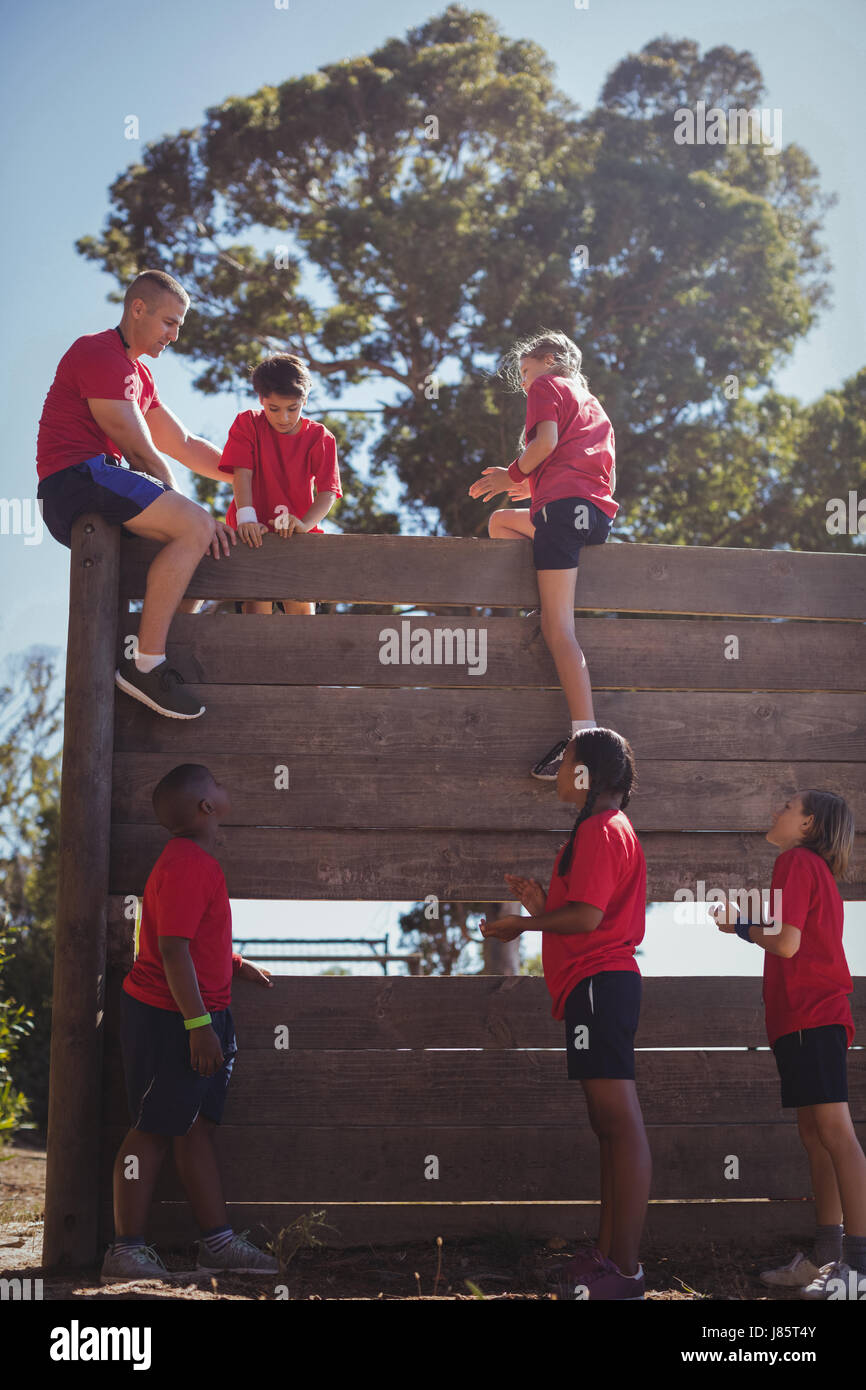Trainer assisting kids to climb a wooden wall during obstacle course ...
