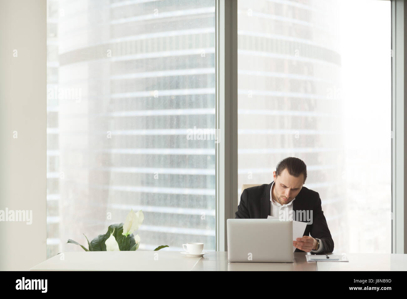 Company leader examines document at desk in modern office with large ...