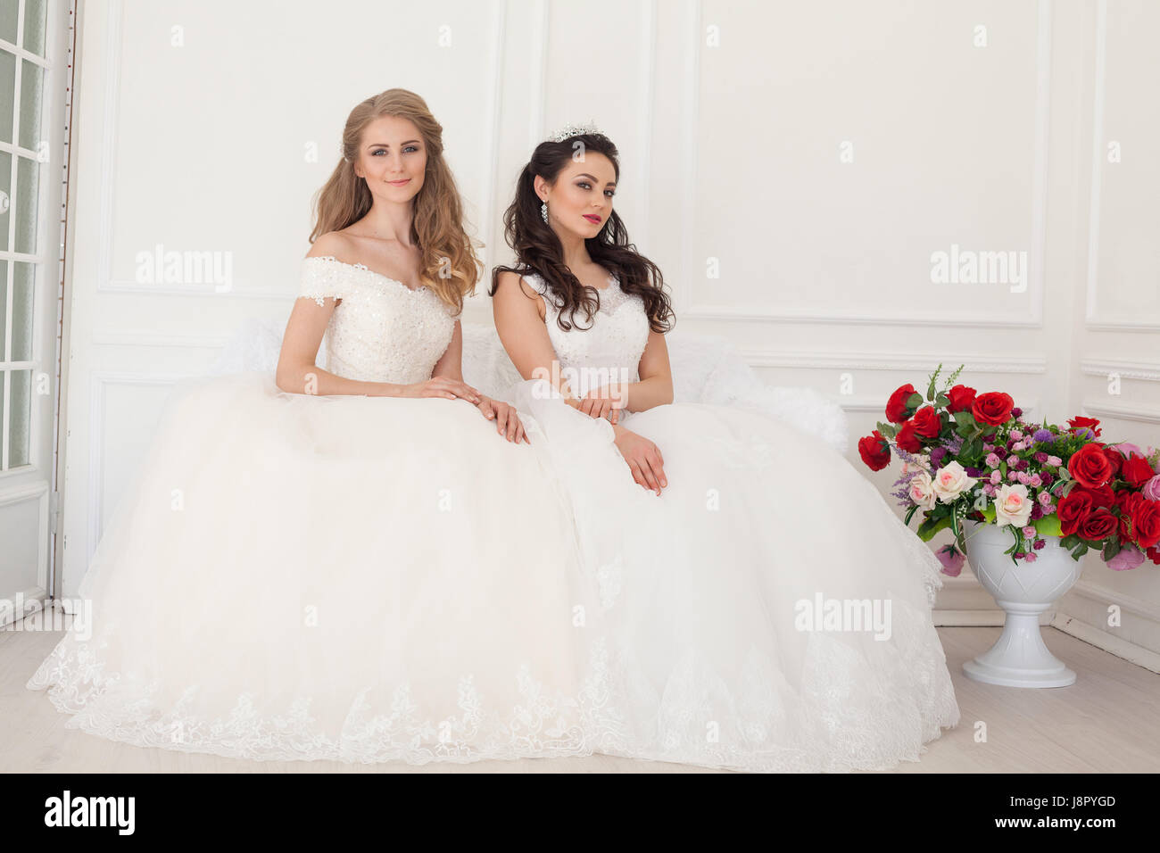 two brides in wedding dresses sit on a white couch Stock Photo - Alamy