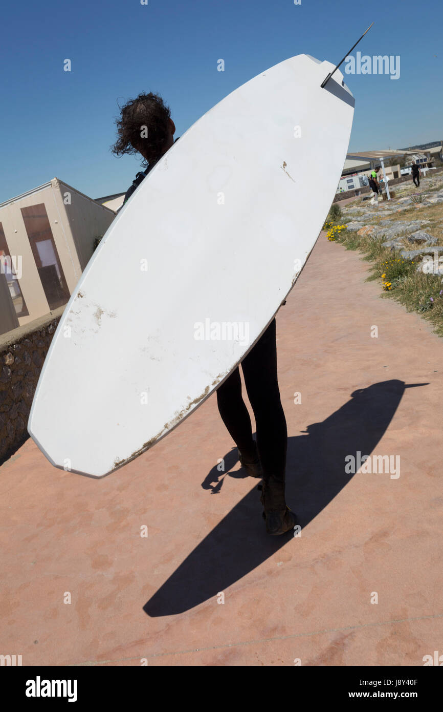 A windsurfer carries his heavy surfboard on his shoulder at the beach ...