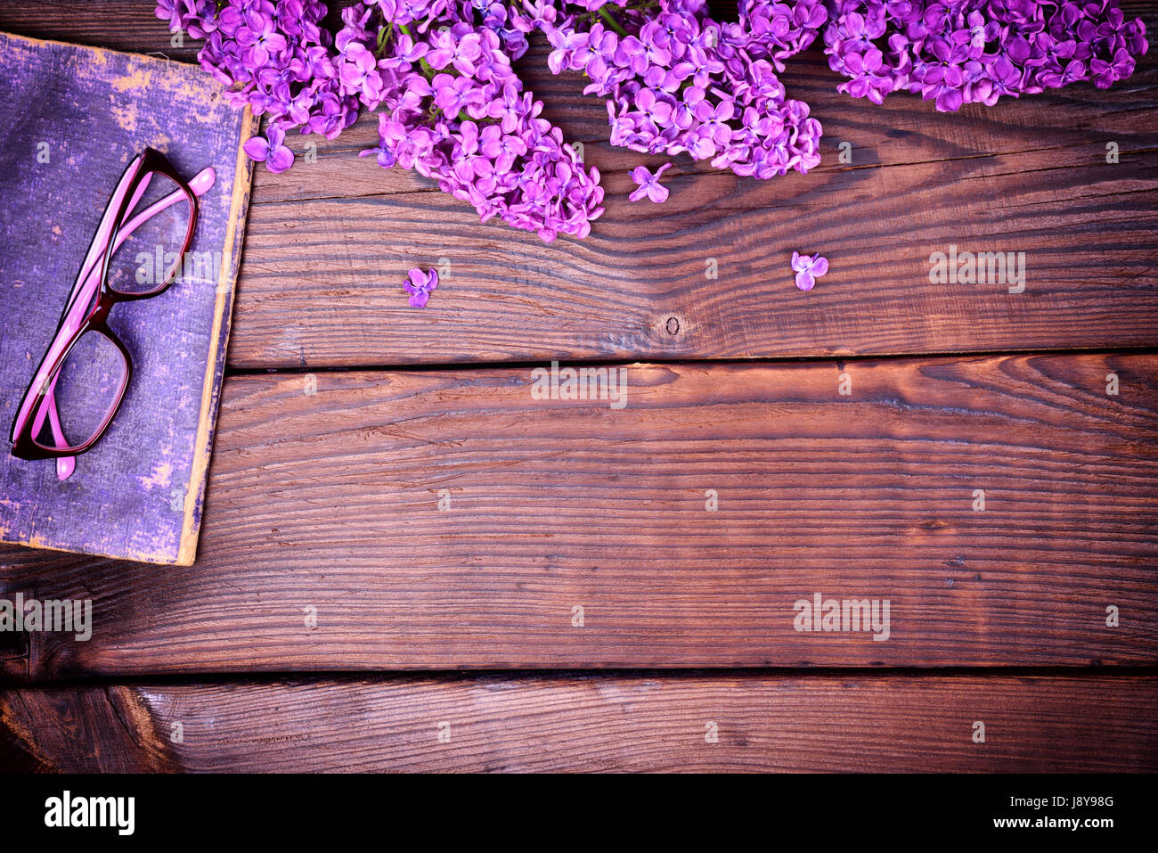 branch of a purple lilac and an old book with glasses on a brown wooden ...