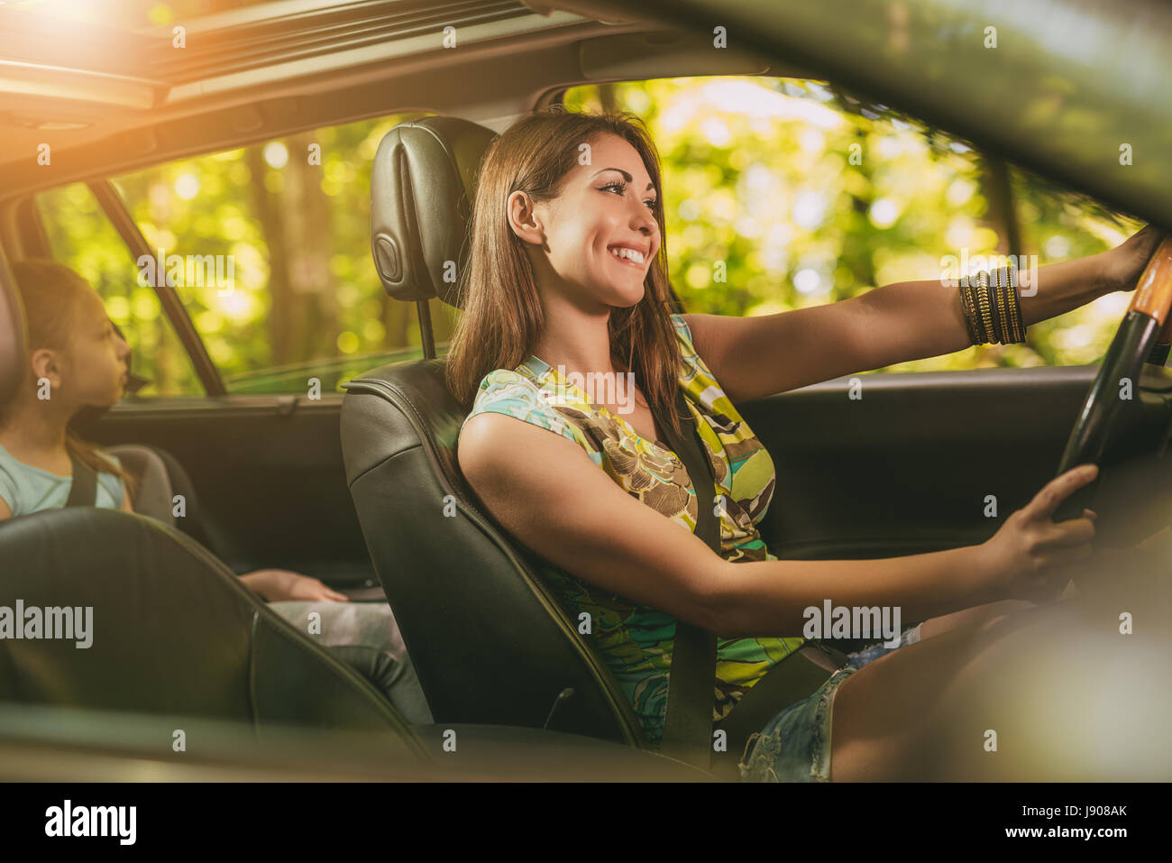 Young beautiful smiling woman driving a car. Her cute daughter sitting ...