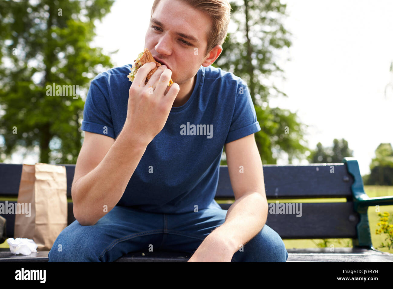 Young Man Sitting On Park Bench Eating Burger Stock Photo - Alamy